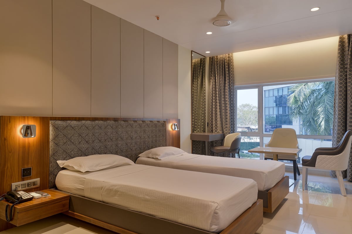 Quarto de hotel com duas camas, uma escrivaninha, uma cadeira e uma janela grande com vista para palmeiras e edifícios.