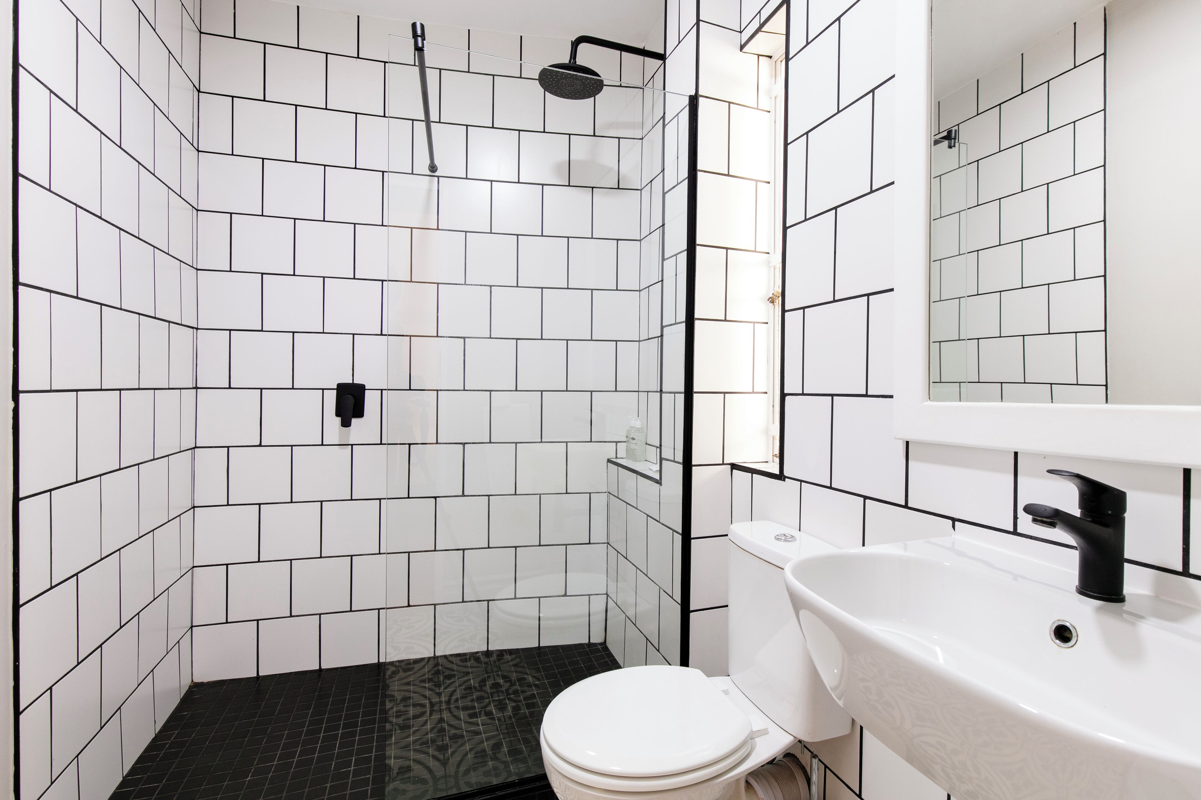 Casa de banho com paredes revestidas de azulejos brancos, área de duche preta, sanita branca e lavatório branco com torneira preta.