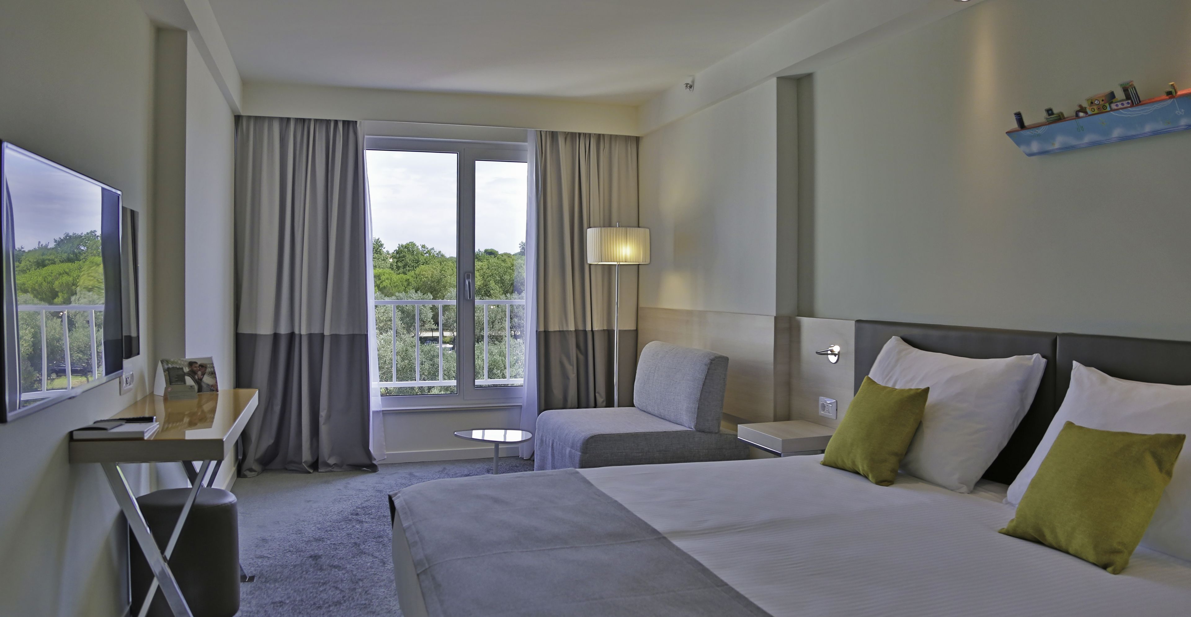 Quarto de hotel com uma cama grande, uma escrivaninha, uma cadeira, uma mesinha e varanda com cortinas.