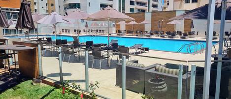 Uma área de piscina no terraço com espreguiçadeiras e guarda-sóis, delimitada por um parapeito de vidro.