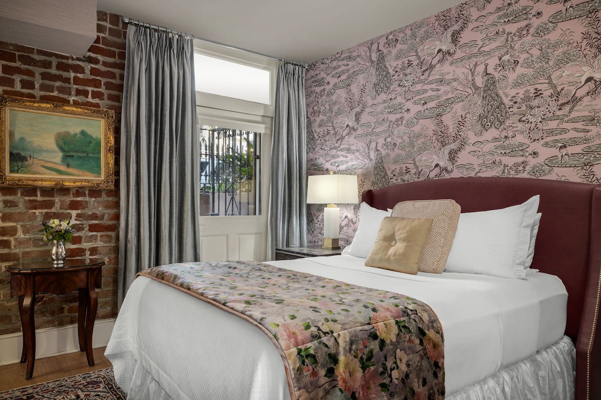 Quarto com uma cama grande, mesa de cabeceira, abajur, janela com cortinas e parede de tijolos com um quadro emoldurado.