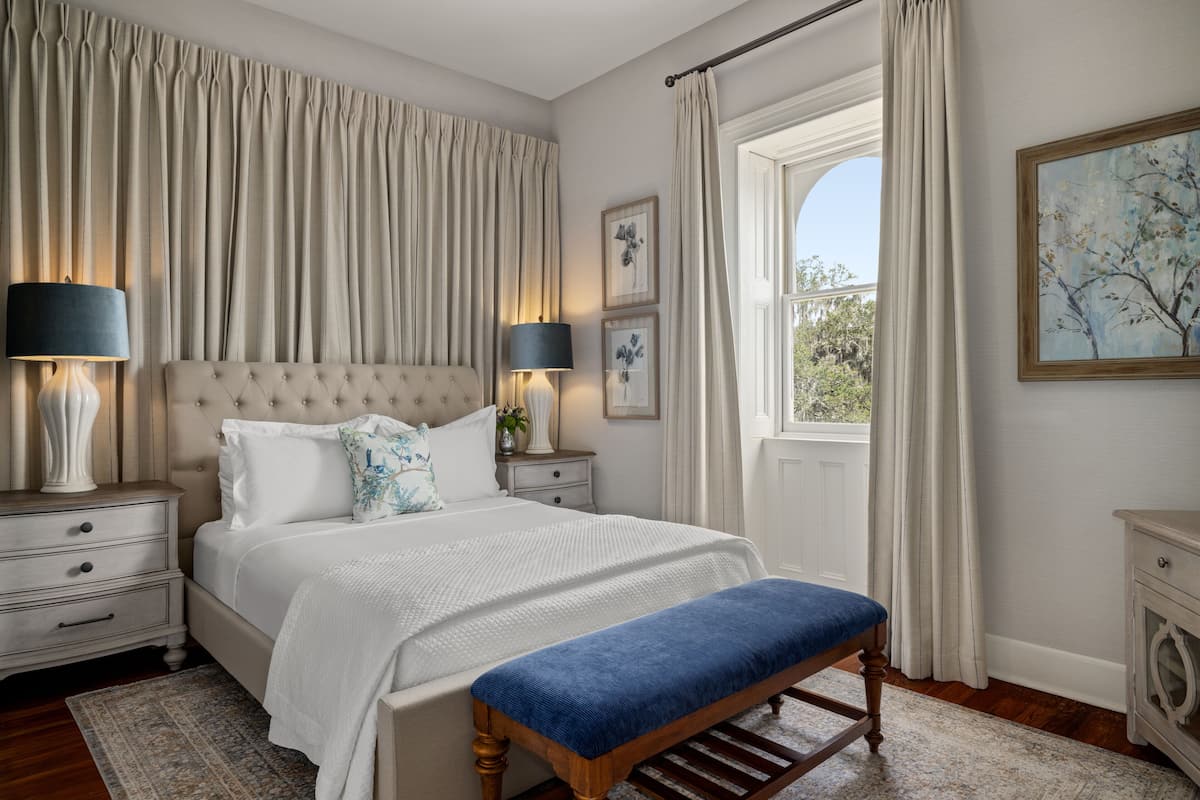 Quarto com cabeceira estofada, cama grande com roupa de cama branca, um banquinho azul, duas mesas de cabeceira com abajures, janela com cortinas e obras de arte emolduradas na parede.