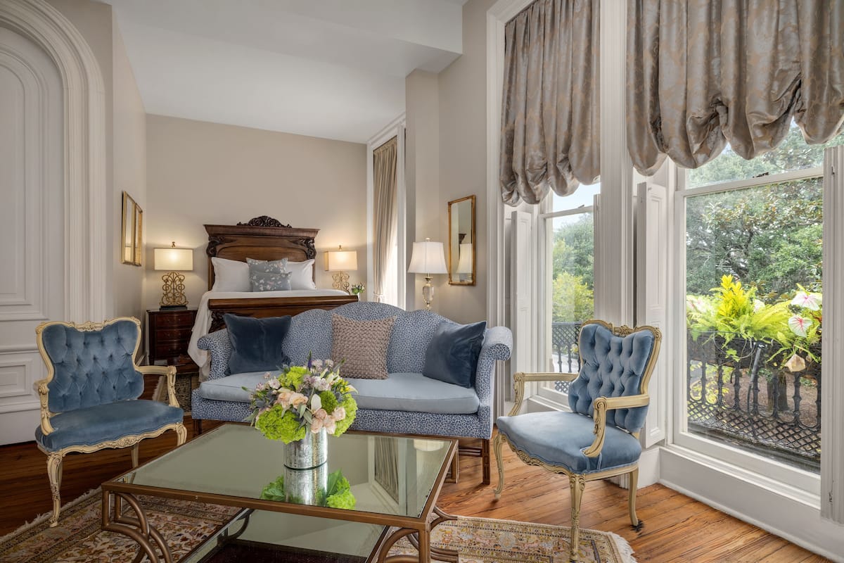 Quarto com cama, duas poltronas azuis estofadas, mesa de centro de vidro com um vaso de flores e vista para o verde através de uma janela.