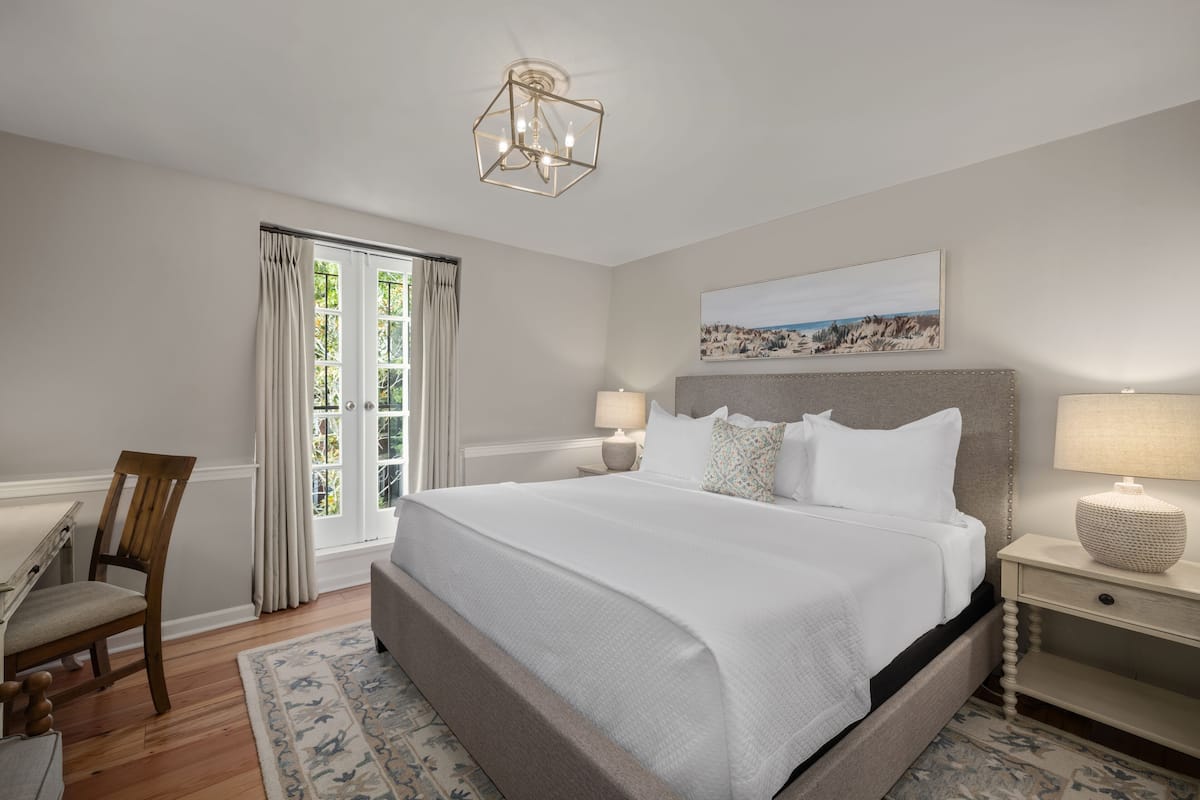 Quarto com uma cama grande, uma cadeira de madeira, uma escrivaninha e vista para o exterior por uma porta de vidro.