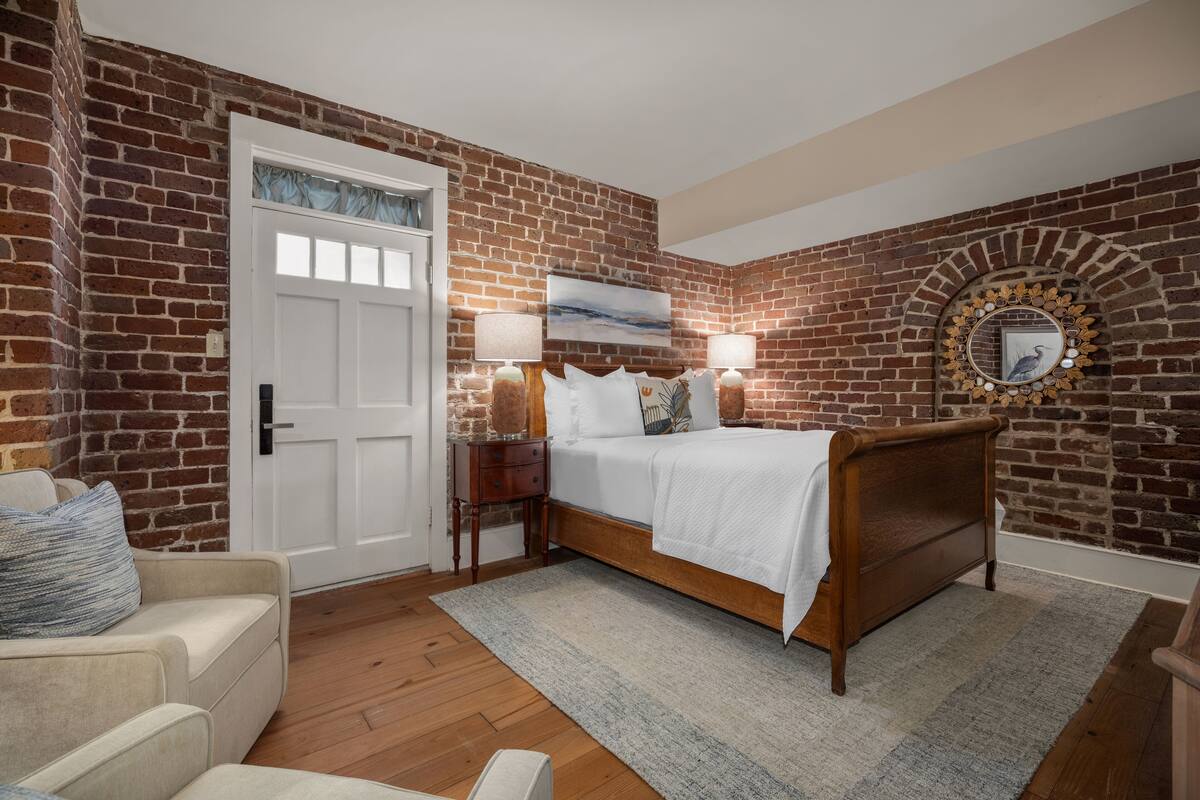Um quarto com parede de tijolos, uma cama de madeira, uma porta branca e uma mesa de cabeceira com luminárias.