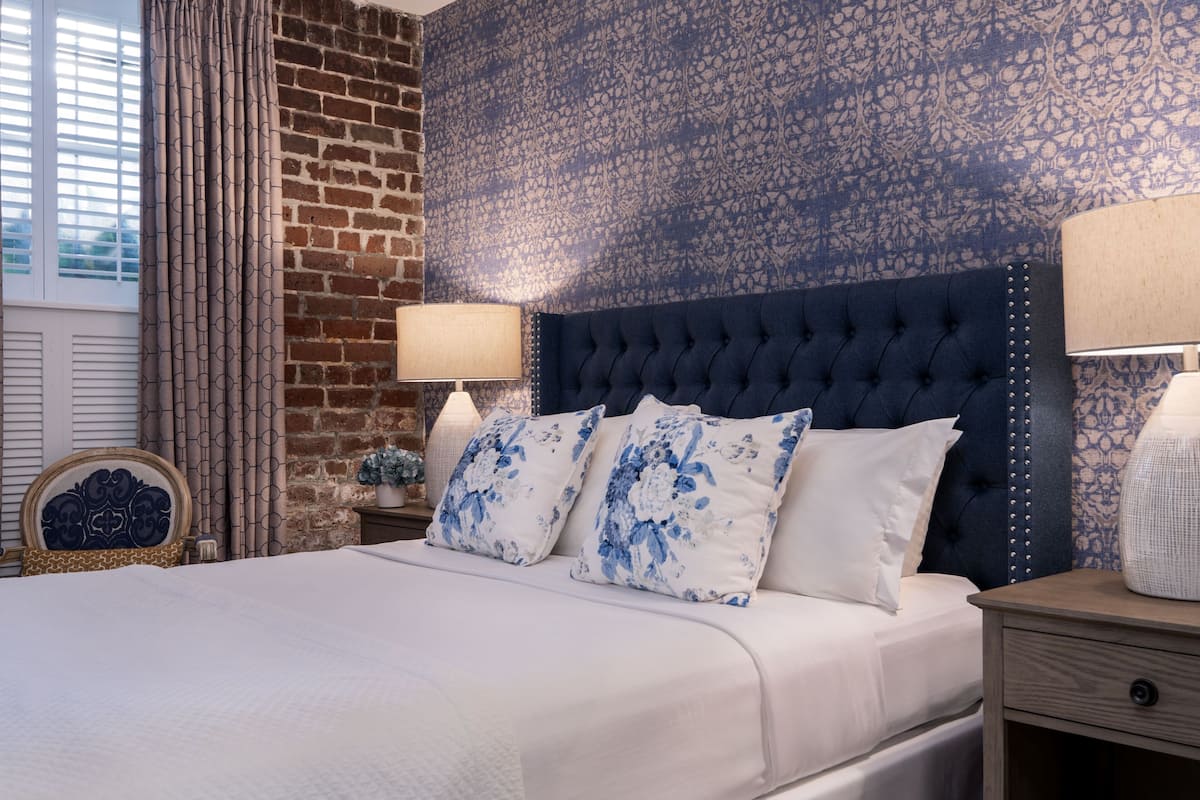 Cama bem arrumada com cabeceira estofada, duas luminárias de apoio e papel de parede com padrão floral.