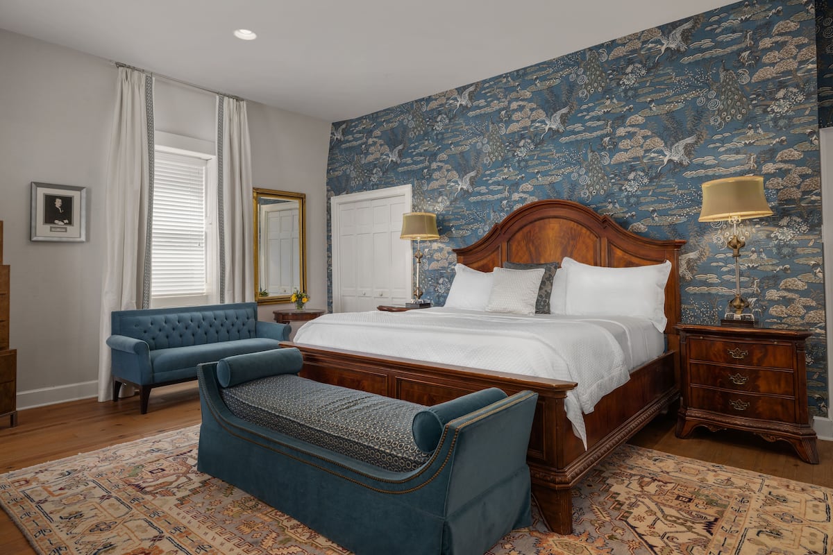 Quarto com cama grande, sofá azul, mesa de cabeceira de madeira e tapete estampado.