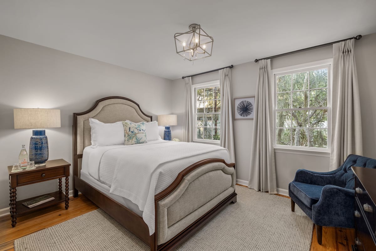 Quarto bem arrumado com uma cama grande, mesa de cabeceira com abajures, uma cadeira e uma janela com cortinas.