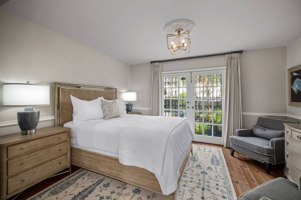 Quarto com cama de madeira, duas mesas de cabeceira com luminárias, uma poltrona cinza e uma janela grande com cortinas.