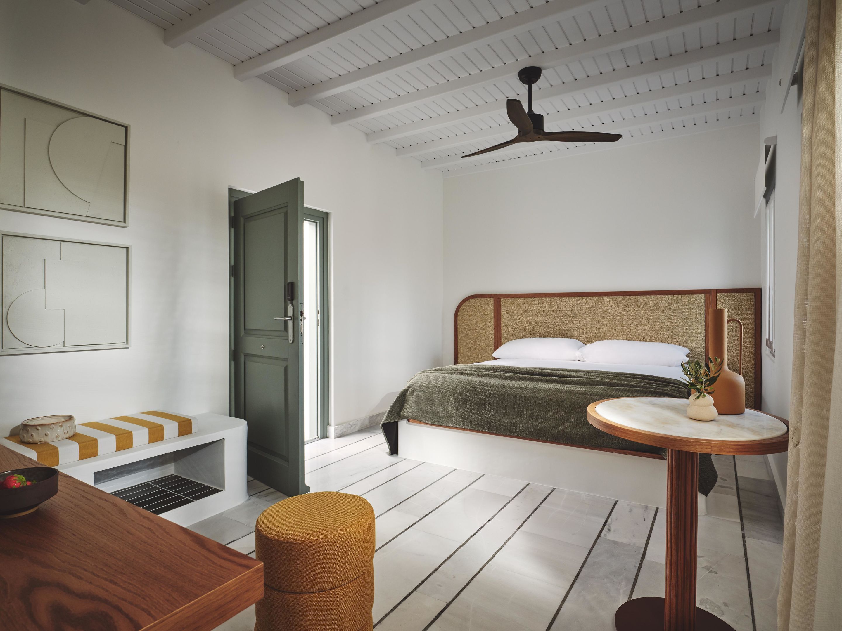 Quarto com cama, um banquinho redondo, uma mesinha pequena e um ventilador de teto, além de uma porta.