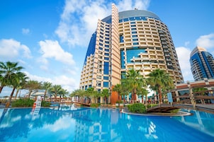 Um hotel moderno com uma piscina grande, palmeiras e um céu azul límpido.