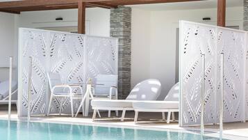Área de piscina com cadeiras de lounge brancas e uma mesa, separada por painéis brancos decorativos com padrões de folhas.