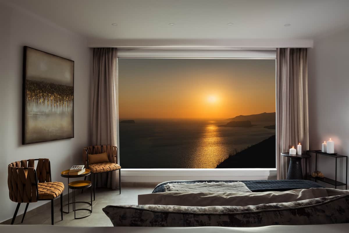 Um quarto moderno com uma janela ampla, uma cama, duas cadeiras, uma mesinha e uma mesa de cabeceira com velas.