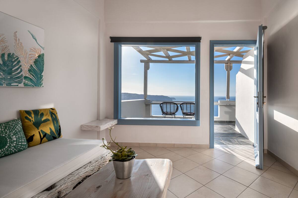 Quarto moderno com uma janela grande, uma cama, uma mesinha com uma planta e vista para o mar.
