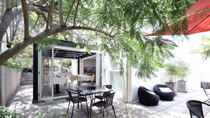 Um pátio externo com mesa e cadeiras, rodeado por plantas em vasos e um guarda-sol vermelho.