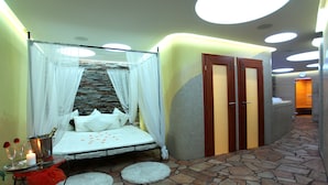 Quarto com piso de pedra, cama com roupa de cama branca, dossel e uma pequena mesa com garrafa e copos.