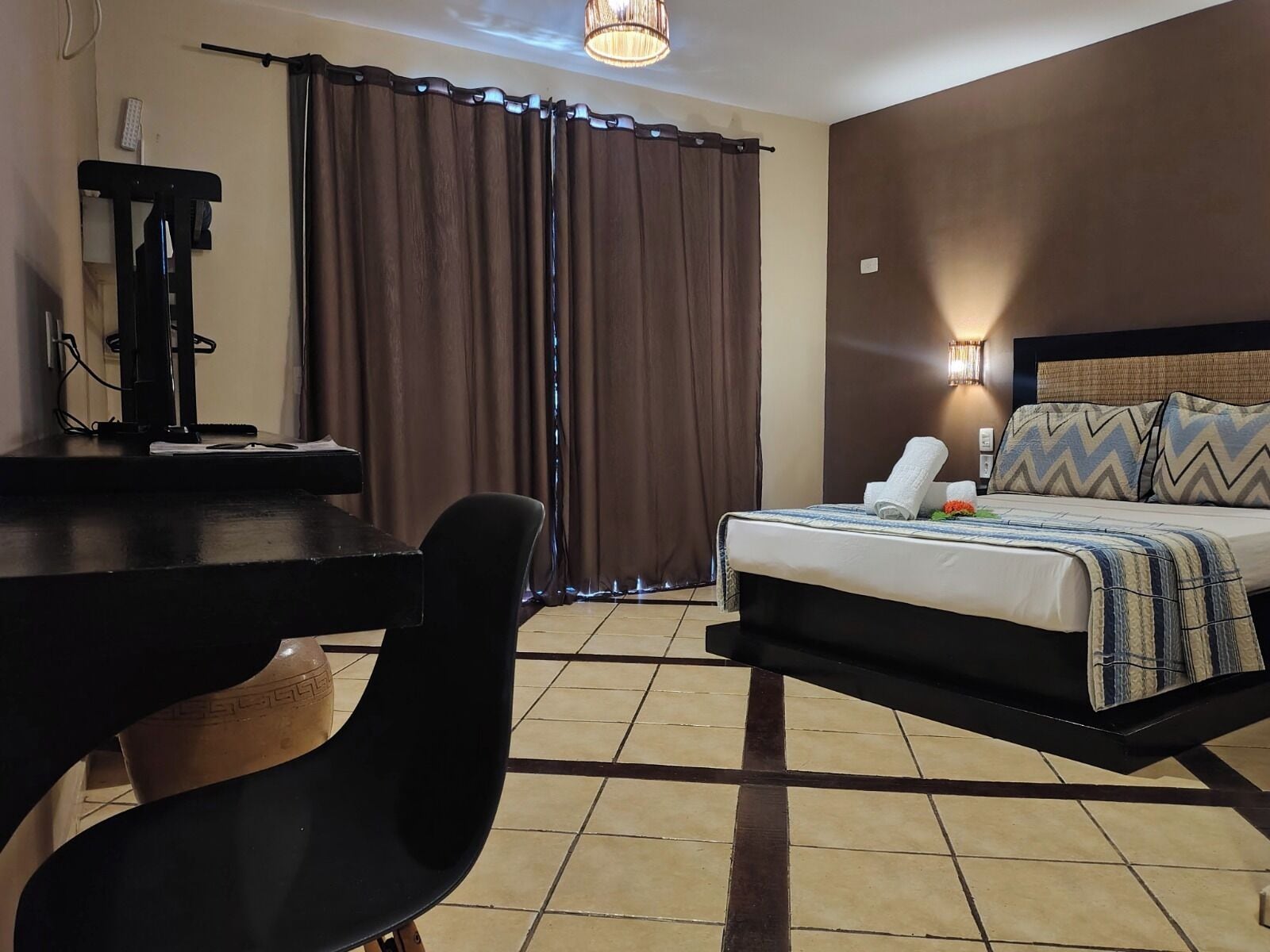 Quarto de hotel com cama, escrivaninha, cadeira e luminária fixa na parede.