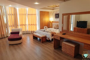 Quarto de hotel com cama, escrivaninha, cadeira, televisão e uma janela grande com cortinas.