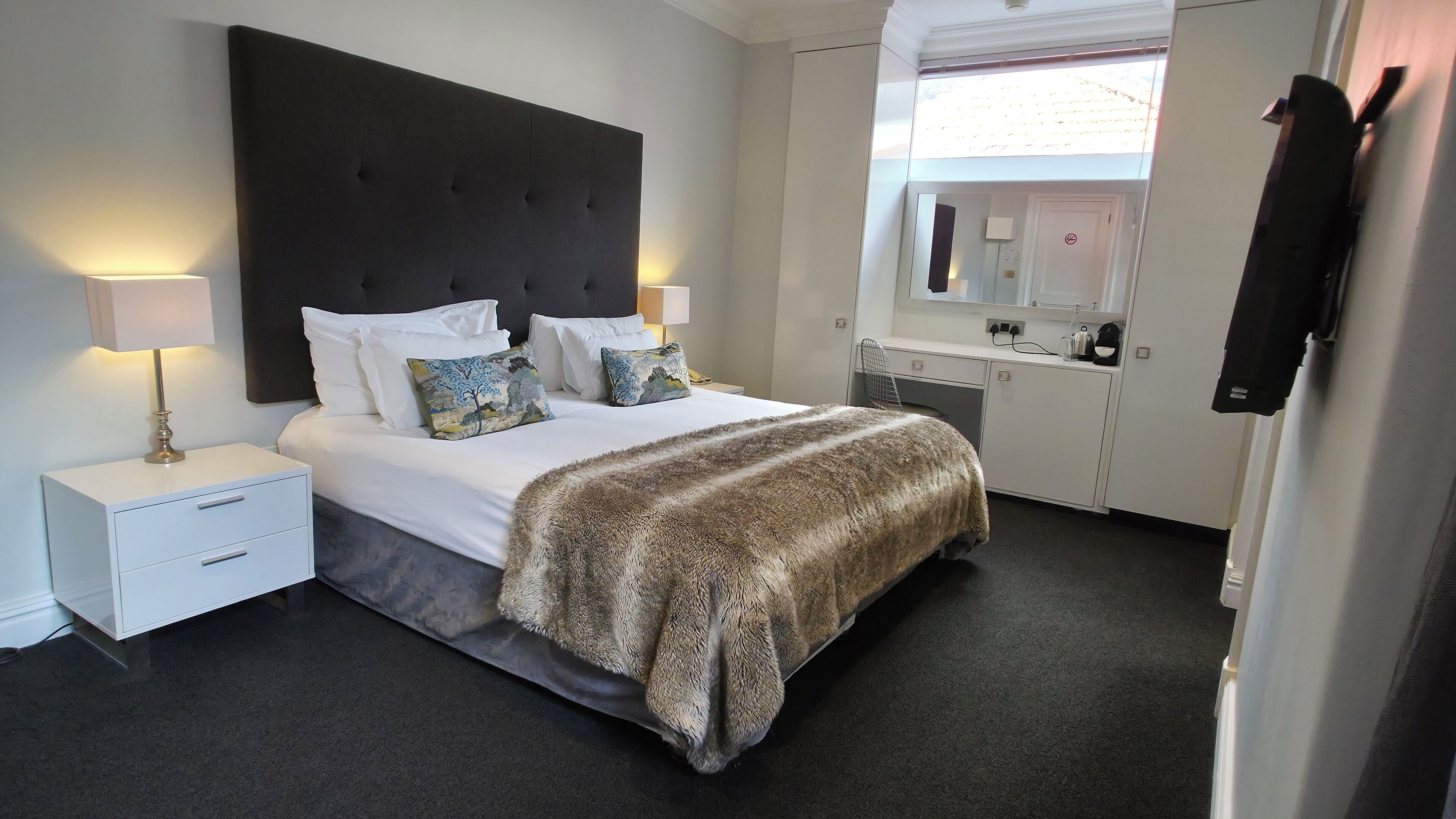 Quarto de hotel com uma cama grande, duas mesas de cabeceira com abajures, um penteado, um espelho e uma janela com persianas.
