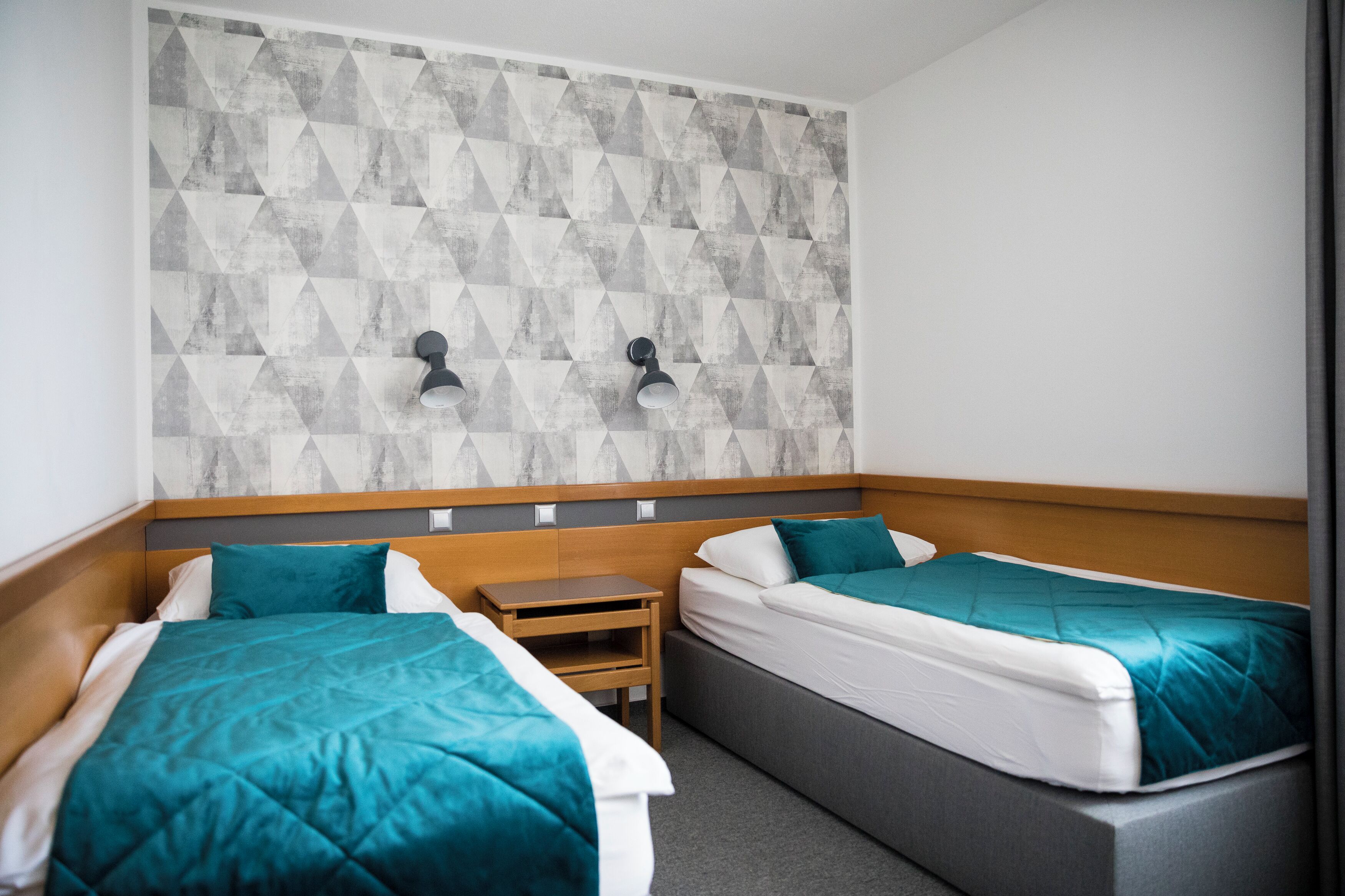 Quarto de hotel com duas camas, cabeceira de madeira, uma mesa pequena e parede com padrão geométrico.