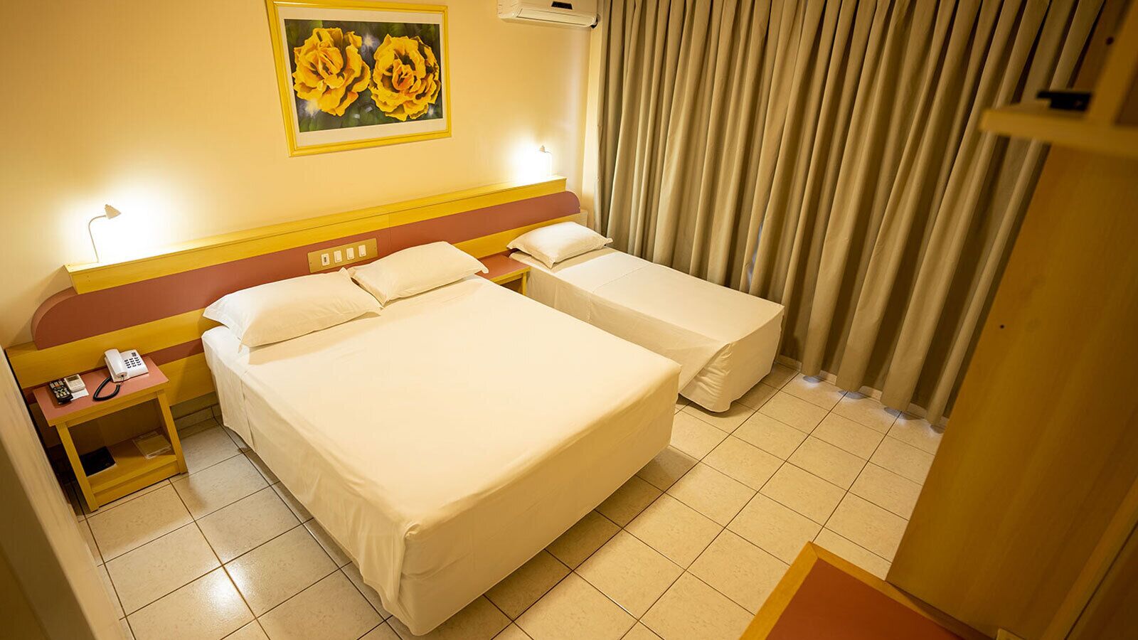 Quarto de hotel com duas camas, uma mesa de cabeceira com telefone, um quadro de flores e uma janela com cortinas.