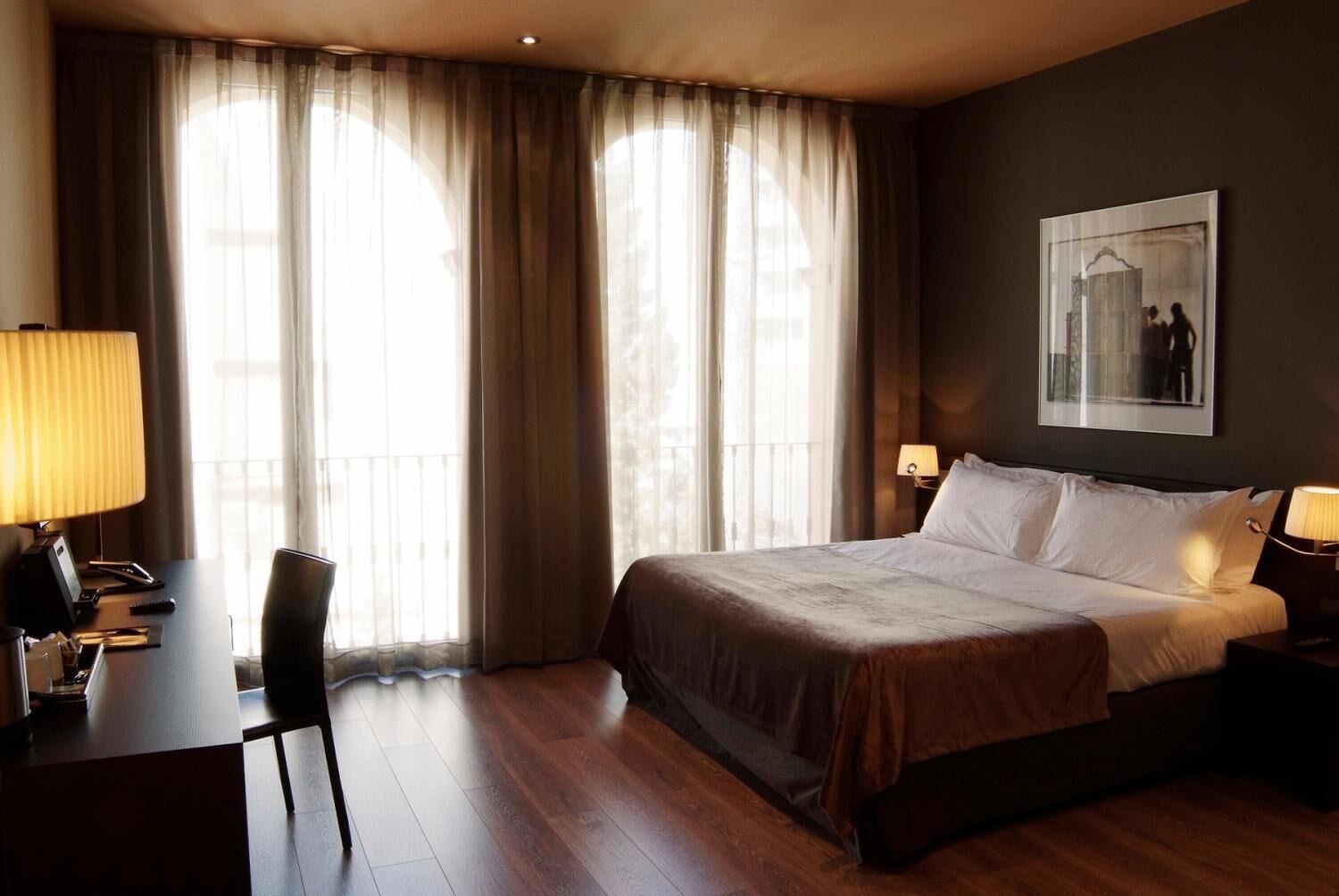 Quarto de hotel com cama, escrivaninha, cadeira e janela com cortinas.
