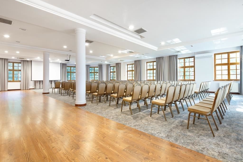 Uma sala de conferências ampla com fileiras de cadeiras, piso de madeira e grandes janelas.