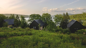 Três cabanas com telhados inclinados, cercadas por vegetação densa e um lago ao fundo.