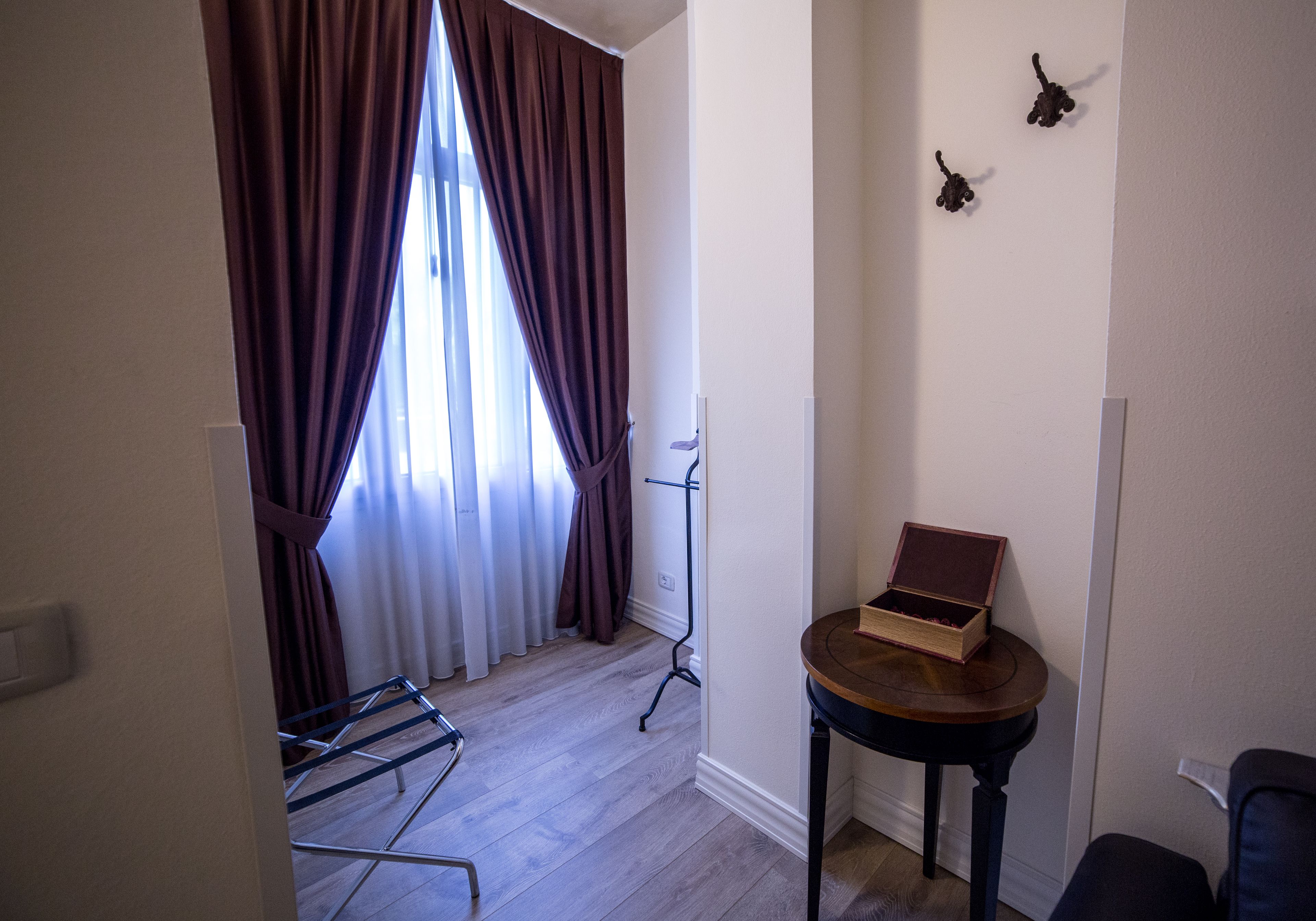 Quarto com piso de madeira, janela com cortinas, pequena mesa com um livro e luminária fixa na parede.