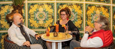 Três pessoas sentadas em uma mesa redonda com bebidas, em um ambiente com papel de parede estampado e um vaso decorativo.