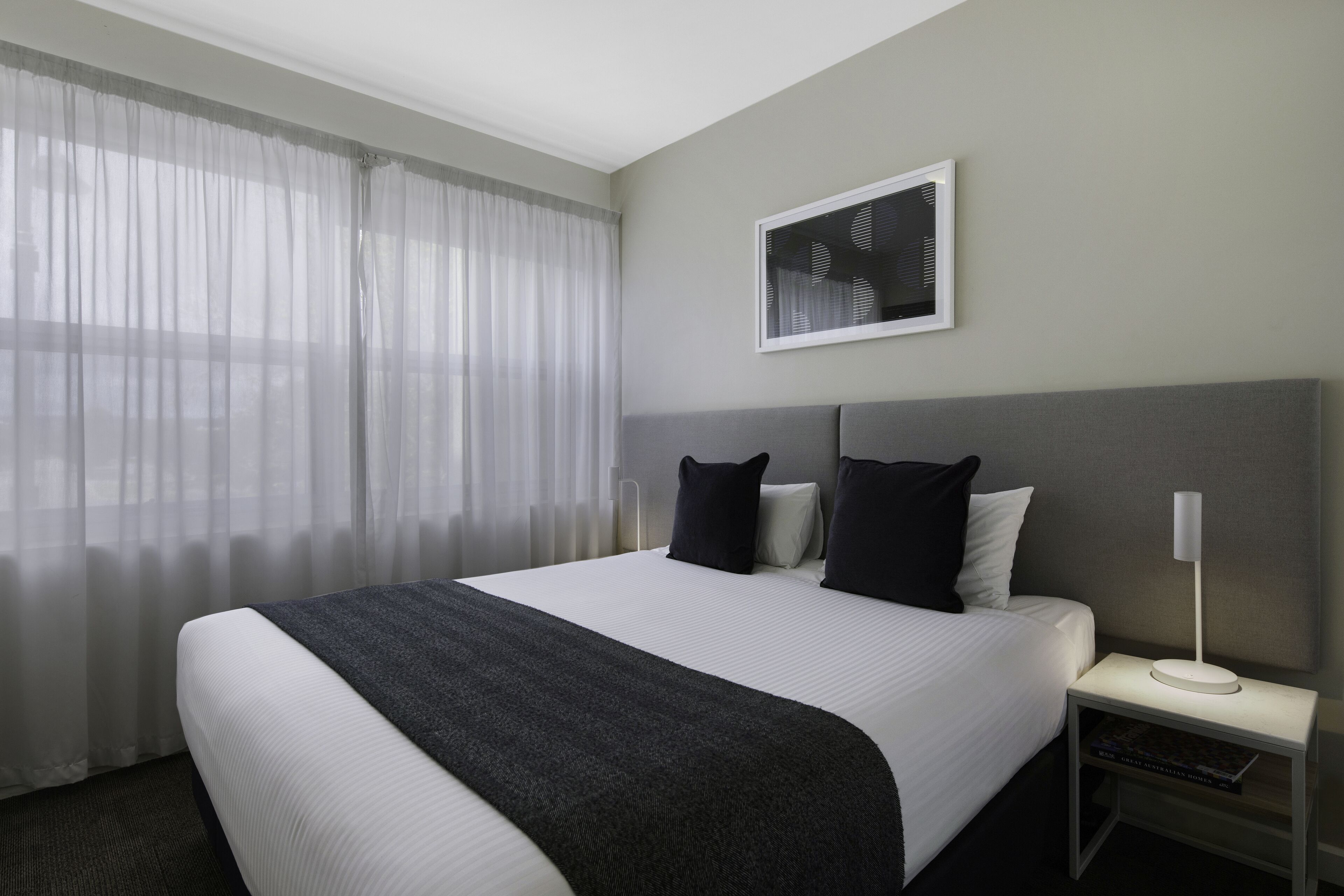 Quarto de hotel com uma cama grande, uma mesa de cabeceira com abajur, uma janela com cortinas leves e um quadro na parede.