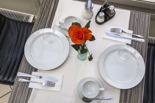Uma mesa de jantar bem posta, com pratos brancos, talheres e uma única rosa laranja.