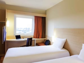 Quarto de hotel com cama, escrivaninha com laptop, cadeira e janela com cortinas.