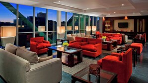 Um lobby de hotel moderno com grandes janelas, assentos em tons de vermelho e bege, e uma TV de tela plana na parede.