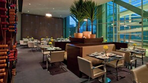 Um restaurante moderno com tetos altos, grandes janelas e uma área central com assentos e uma planta decorativa.