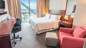Quarto de hotel com uma cama grande, uma escrivaninha, uma cadeira, um sofá vermelho e vista para a cidade através de uma janela ampla.