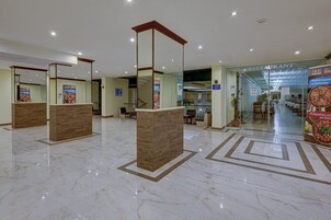 Um saguão de hotel com piso de mármore, balcões de recepção e um restaurante ao fundo.