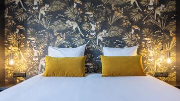 Quarto de hotel com uma cama, duas almofadas amarelas e papel de parede com tema de animais e plantas tropicais.