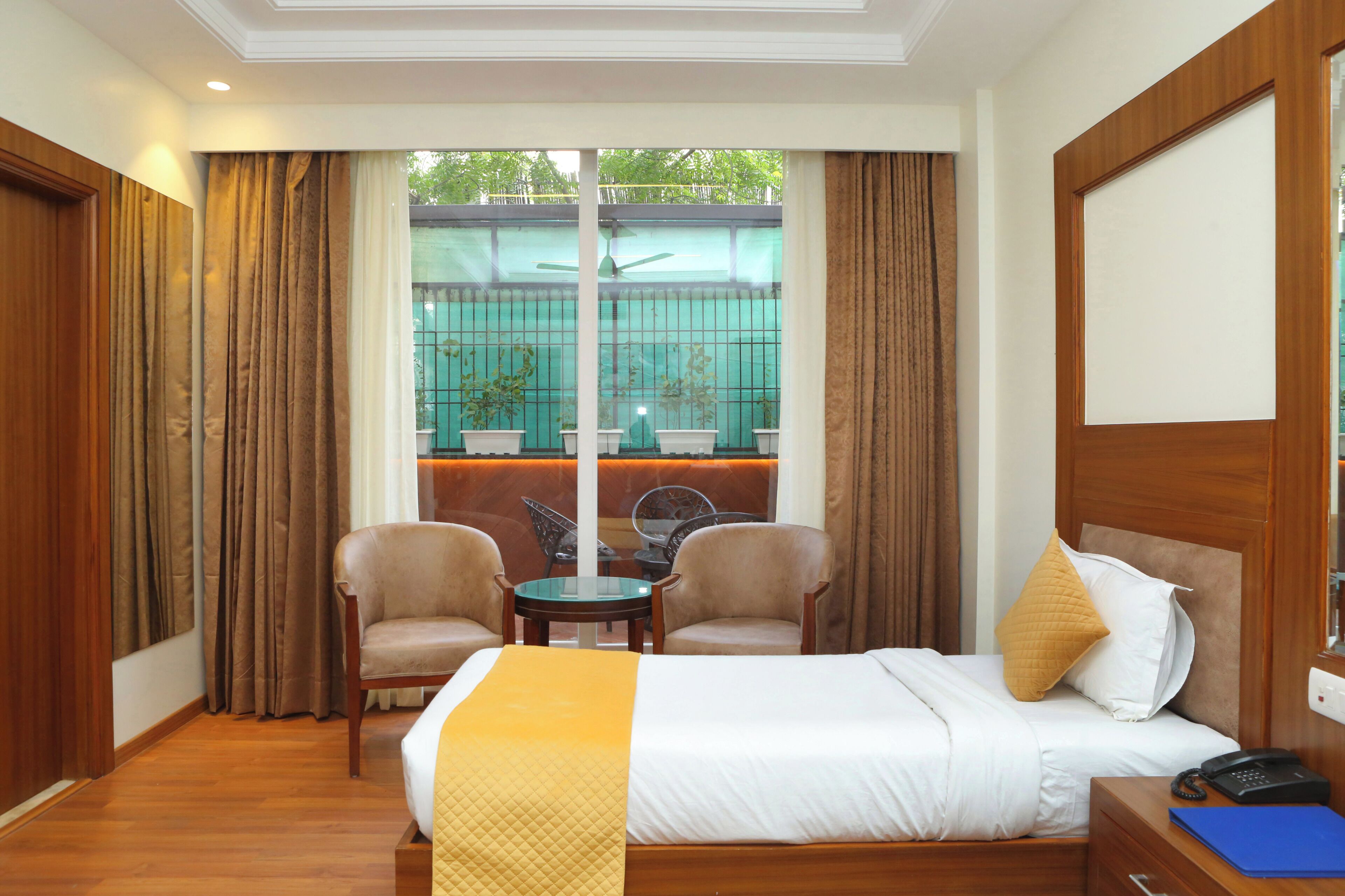 Quarto de hotel com uma cama, duas poltronas, uma mesinha e vista para uma área verde externa.