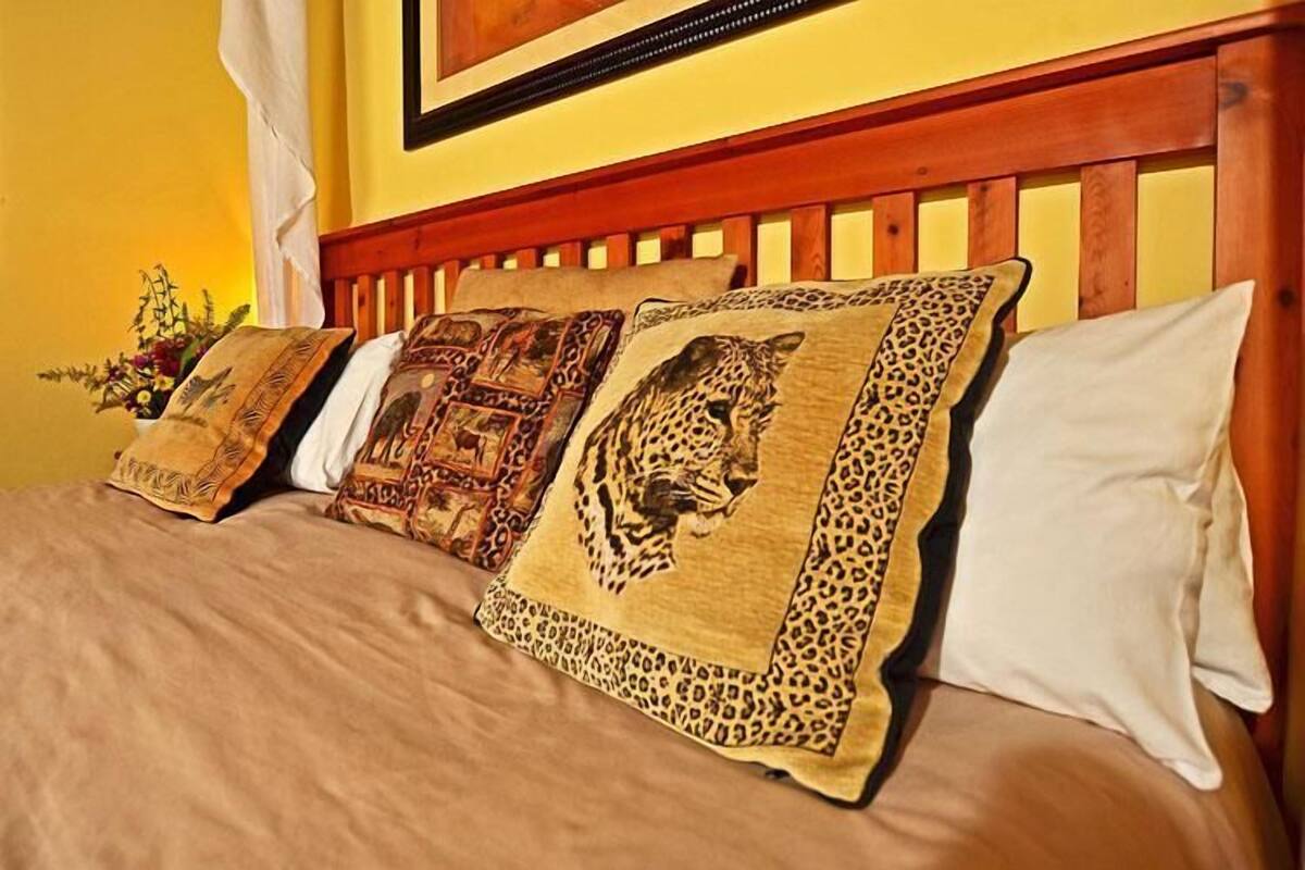Uma cama com cabeceira de madeira, várias almofadas com estampas de animais e um vaso com flores do lado esquerdo.