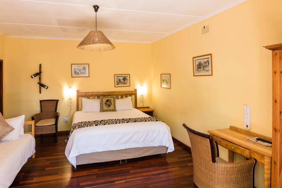 Quarto de hotel com uma cama grande, duas camas menores, uma escrivaninha de madeira, cadeiras de vime e quadros nas paredes.
