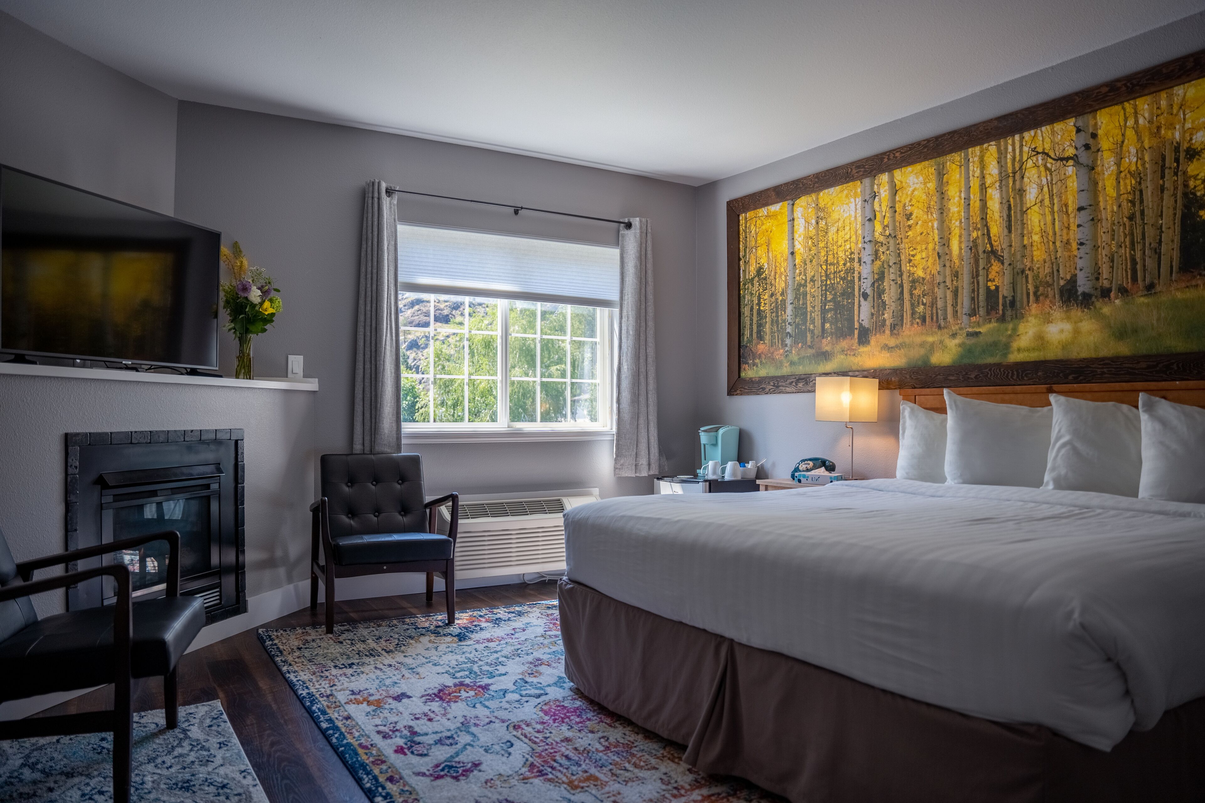 Quarto de hotel com uma cama grande, televisão na parede, lareira, uma poltrona, janela com cortinas e um quadro de uma floresta.
