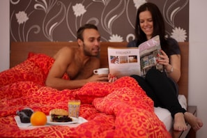 Um casal em uma cama, com uma revista, uma xícara e um prato com comida e bebida.