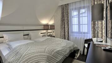 Quarto de hotel com uma cama grande, uma escrivaninha, uma cadeira e uma janela com cortinas.