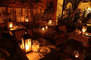 Restaurante com iluminação suave, mesas preparadas para refeições, cercado por plantas em vasos e iluminação acolhedora.