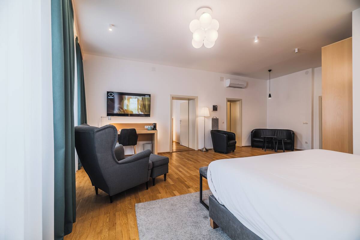 Quarto de hotel moderno com uma cama grande, uma escrivaninha com cadeira, um sofá e uma TV fixada na parede.