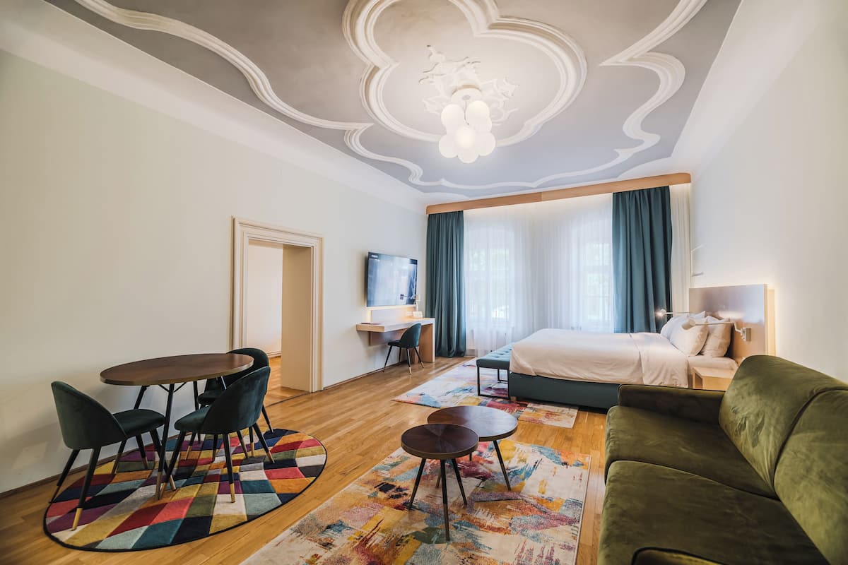 Quarto de hotel com cama, escrivaninha, televisão, sofá verde e tapete colorido.