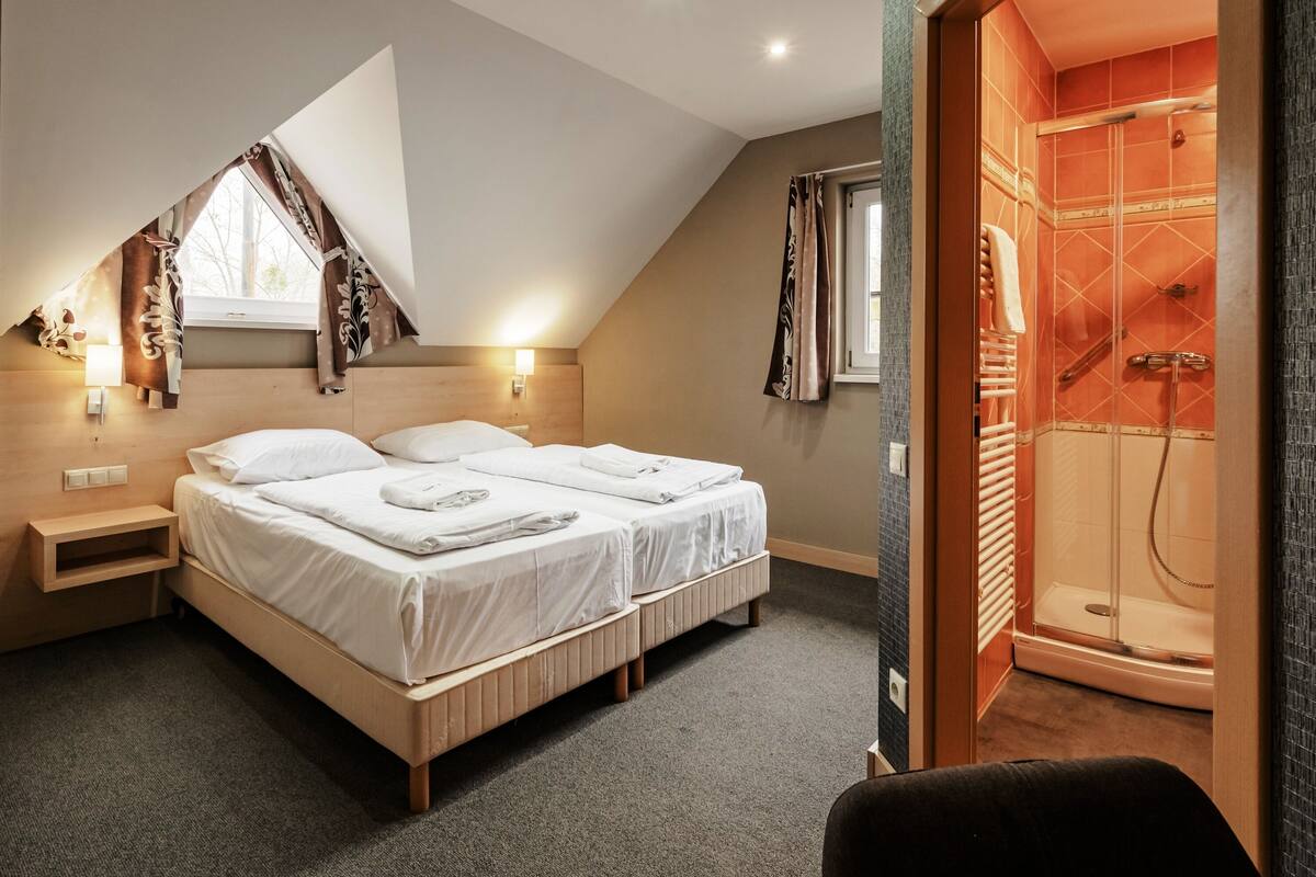 Quarto com duas camas, janela com cortinas, mesa de cabeceira e banheiro com porta de vidro.