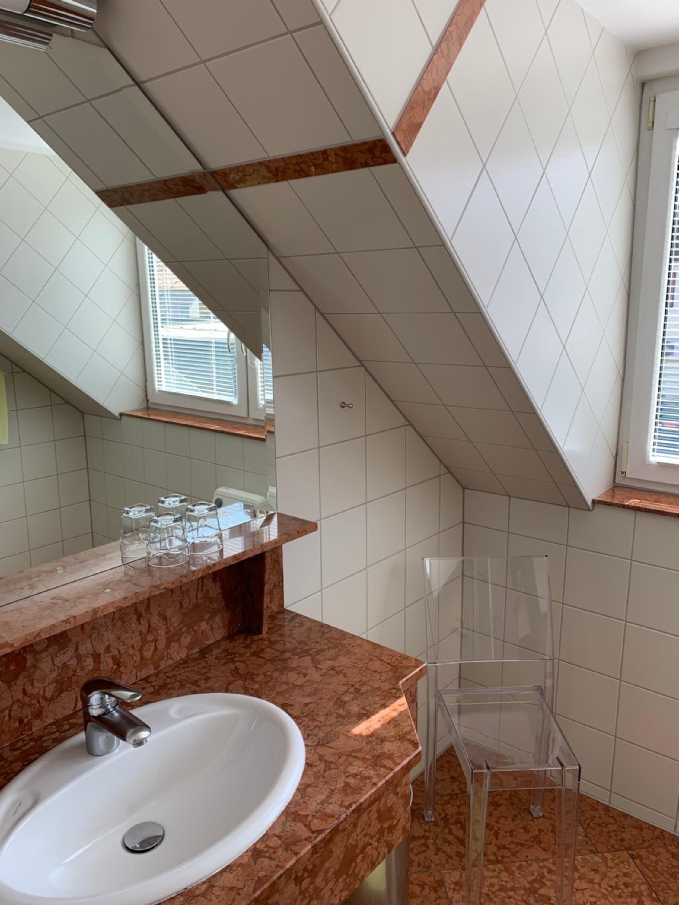 Um banheiro com pia branca, bancada de mármore, box de vidro transparente e janela com persianas.