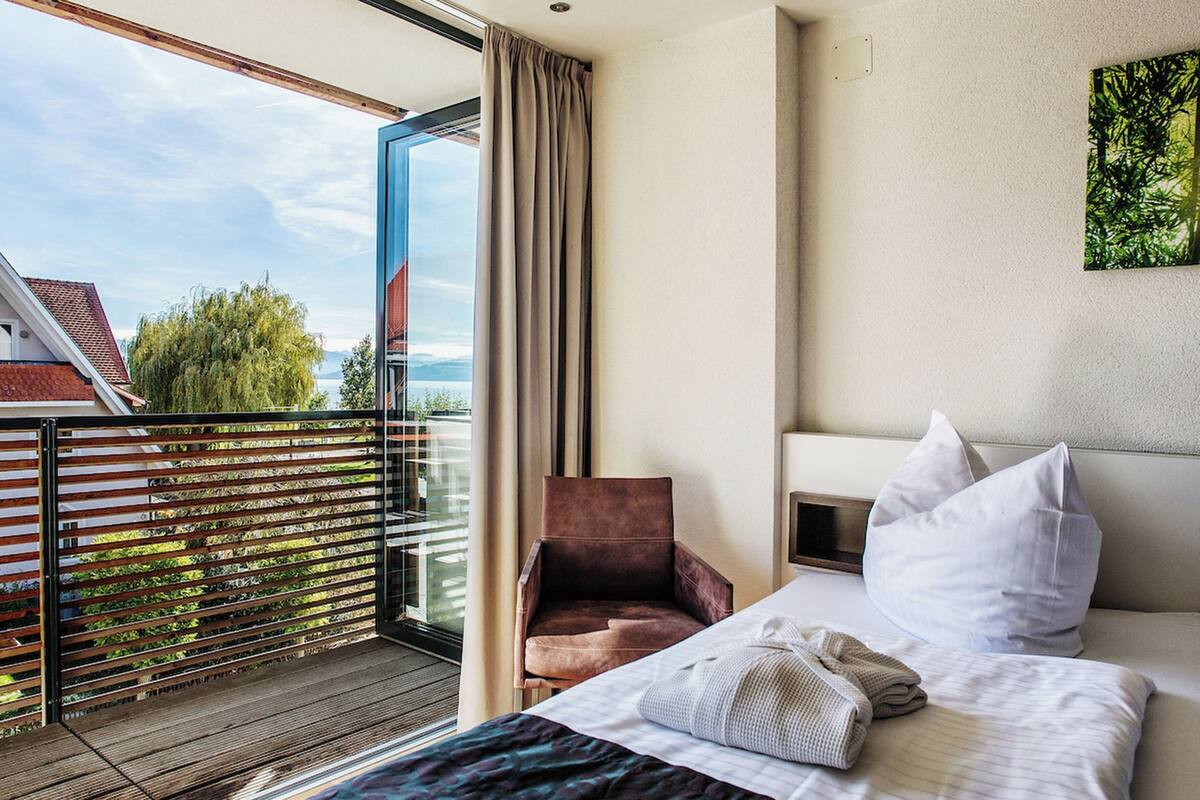 Quarto de hotel moderno com uma janela grande, varanda, cama com lençóis brancos e uma poltrona marrom.